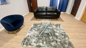 Uma sala de estar com um sofá de couro preto, uma poltrona azul e uma mesa de centro de vidro sobre um tapete estampado.