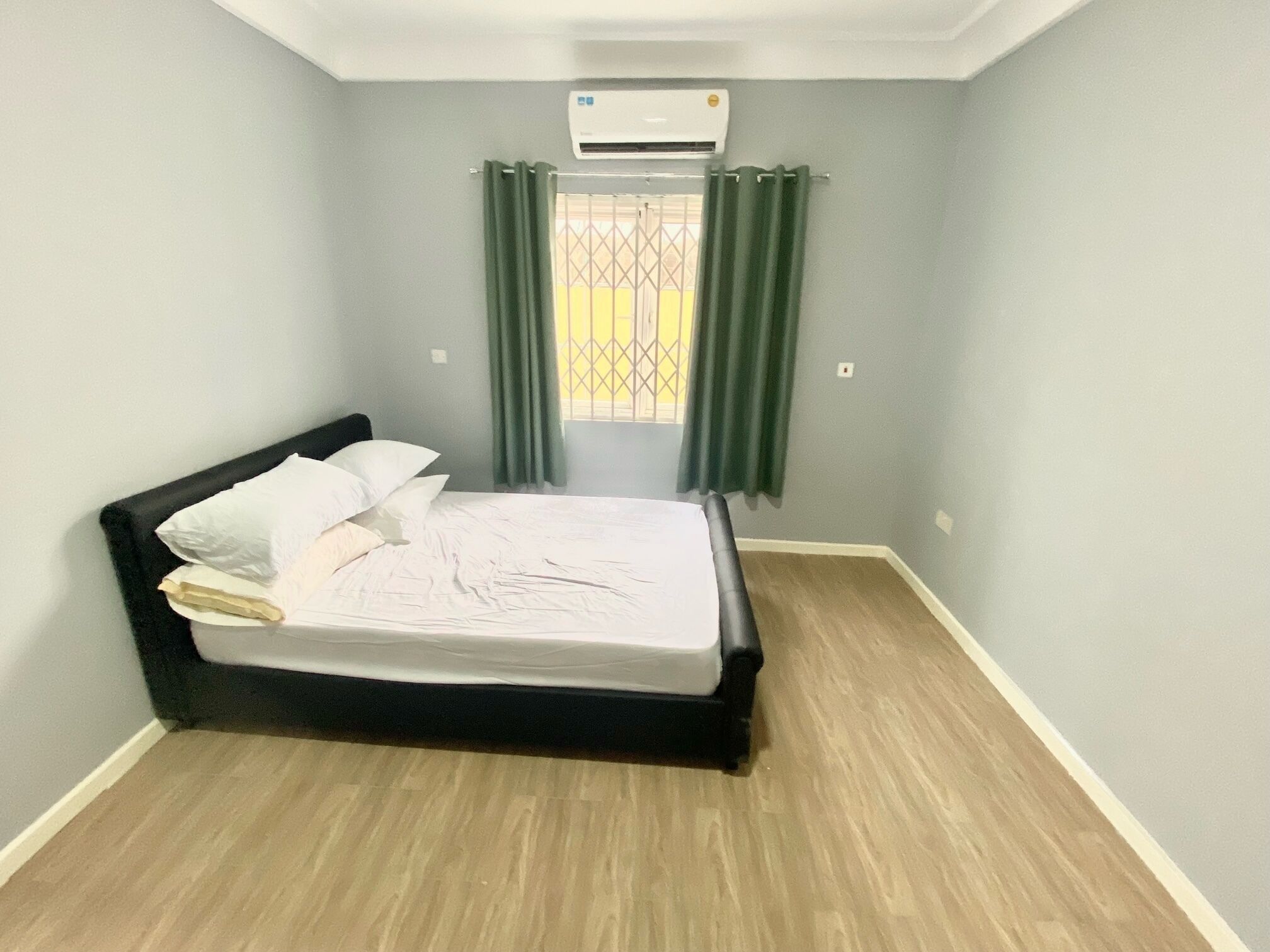 Quarto com uma cama, janela com cortinas, aparelho de ar condicionado e piso de madeira.