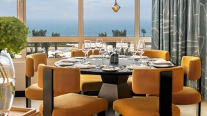 Uma sala de jantar com uma mesa posta para seis pessoas, janelas amplas com vista para o mar e luminárias pendentes.