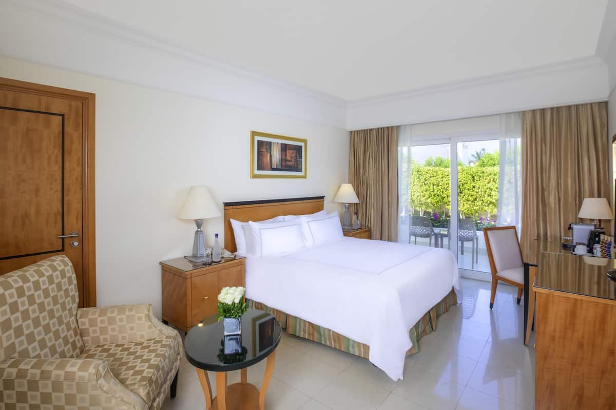Quarto de hotel com cama, sofá, uma mesinha com um vaso de flores, mesa de cabeceira com abajur e vista para o verde pela janela.