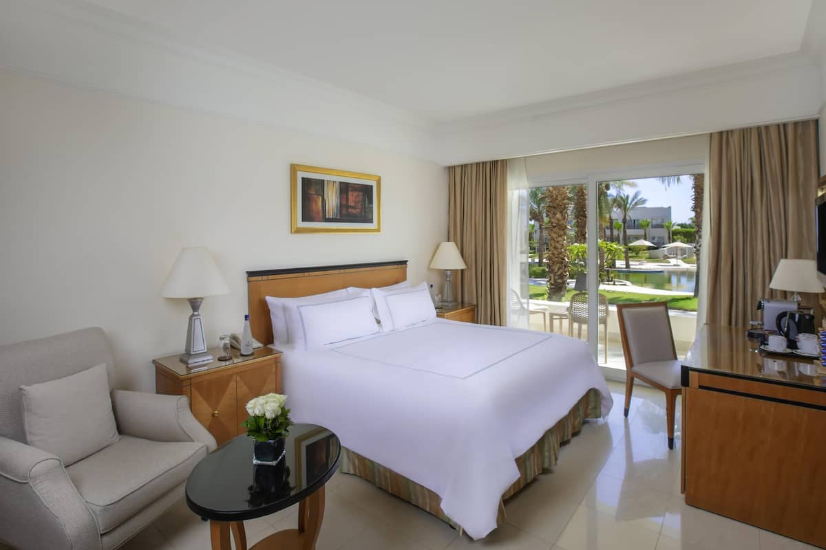 Quarto de hotel com cama, sofá, escrivaninha e vista para uma piscina e palmeiras.
