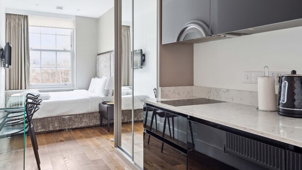 Quarto de hotel moderno com cama, mesa de vidro, área de cozinha com pia e vista para a cidade pela janela.