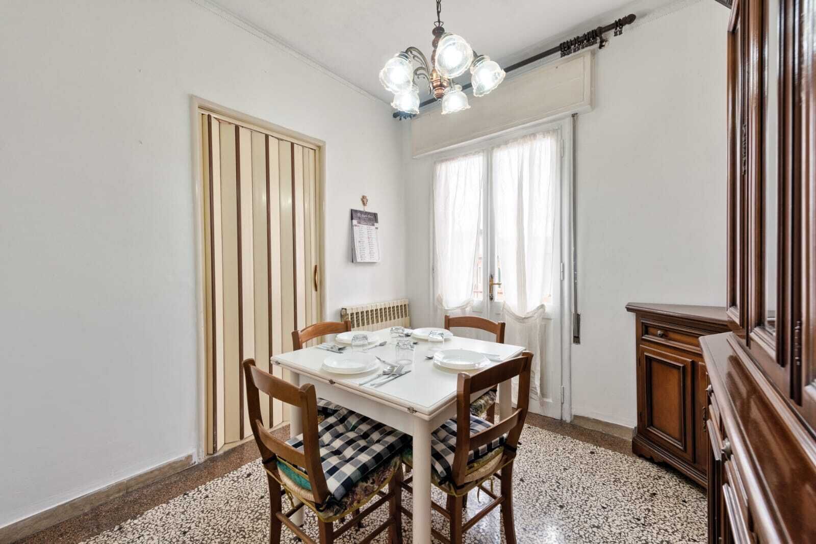 Uma sala de jantar com uma mesa branca posta para quatro pessoas, cadeiras de madeira com almofadas xadrez, um armário de madeira e uma janela com cortinas leves.