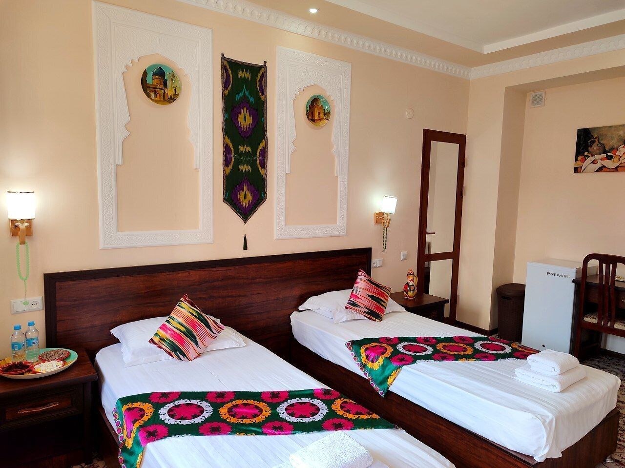 Quarto de hotel com duas camas de solteiro, cada uma com um cobertor colorido e um travesseiro. Há luminárias fixadas na parede e uma pequena mesa com uma garrafa e copos.