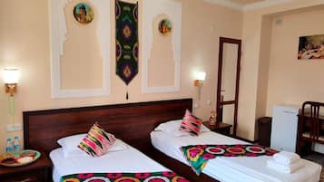 Quarto de hotel com duas camas de solteiro, cada uma com um cobertor colorido e um travesseiro. Há luminárias fixadas na parede e uma pequena mesa com uma garrafa e copos.