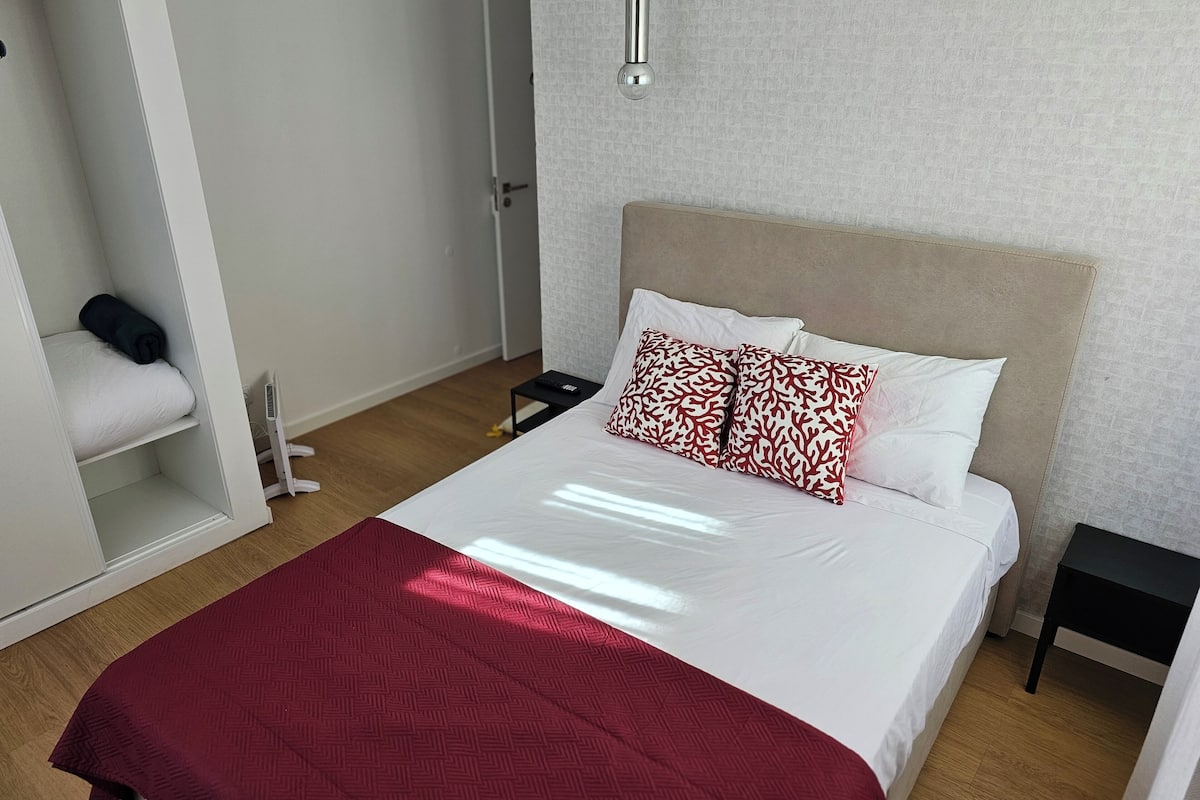 Cama bem arrumada com lençóis brancos e duas almofadas estampadas, mesa de cabeceira com controle remoto e luminária pendente sobre a cama.