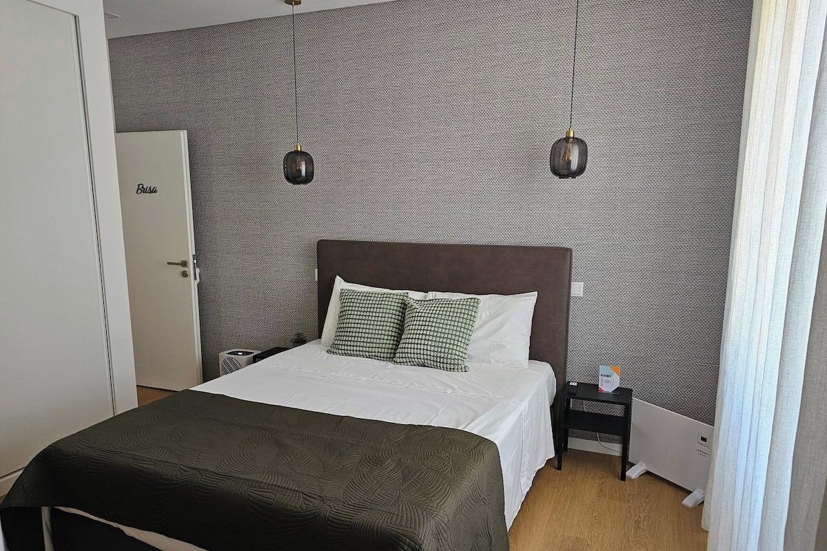 Quarto bem arrumado com cama, mesa de cabeceira, aquecedor fixado na parede e uma porta.