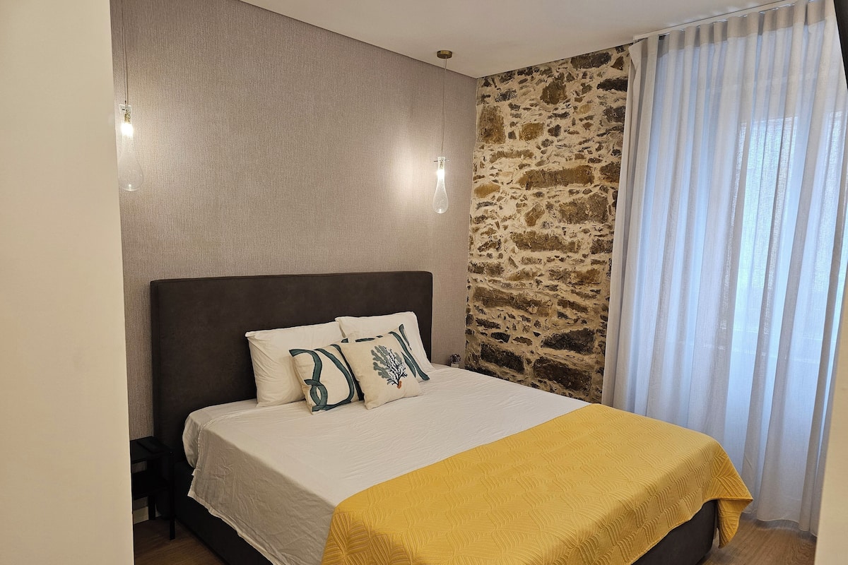 Quarto com parede de pedra, cama com colcha branca e manta amarela, e mesa de cabeceira com abajur.