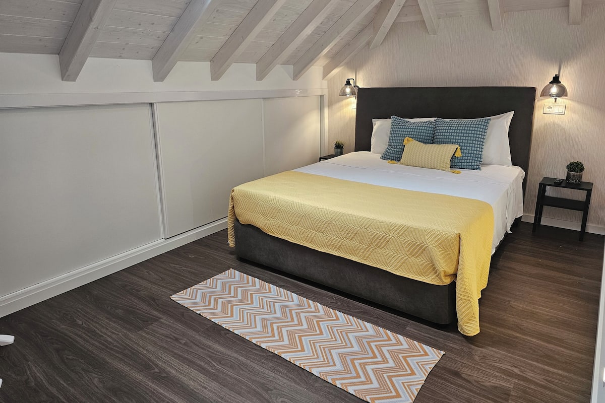 Quarto com cama, mesa de cabeceira, luminária, planta em vaso e tapete com padrão em chevron.