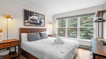 Quarto bem arrumado com uma cama grande, mesas de cabeceira, escrivaninha e janela com vista para uma floresta.