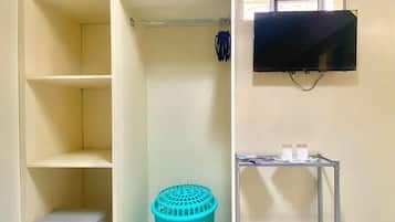 Quarto com televisão na parede, cesto de roupa e uma pequena mesa com objetos.