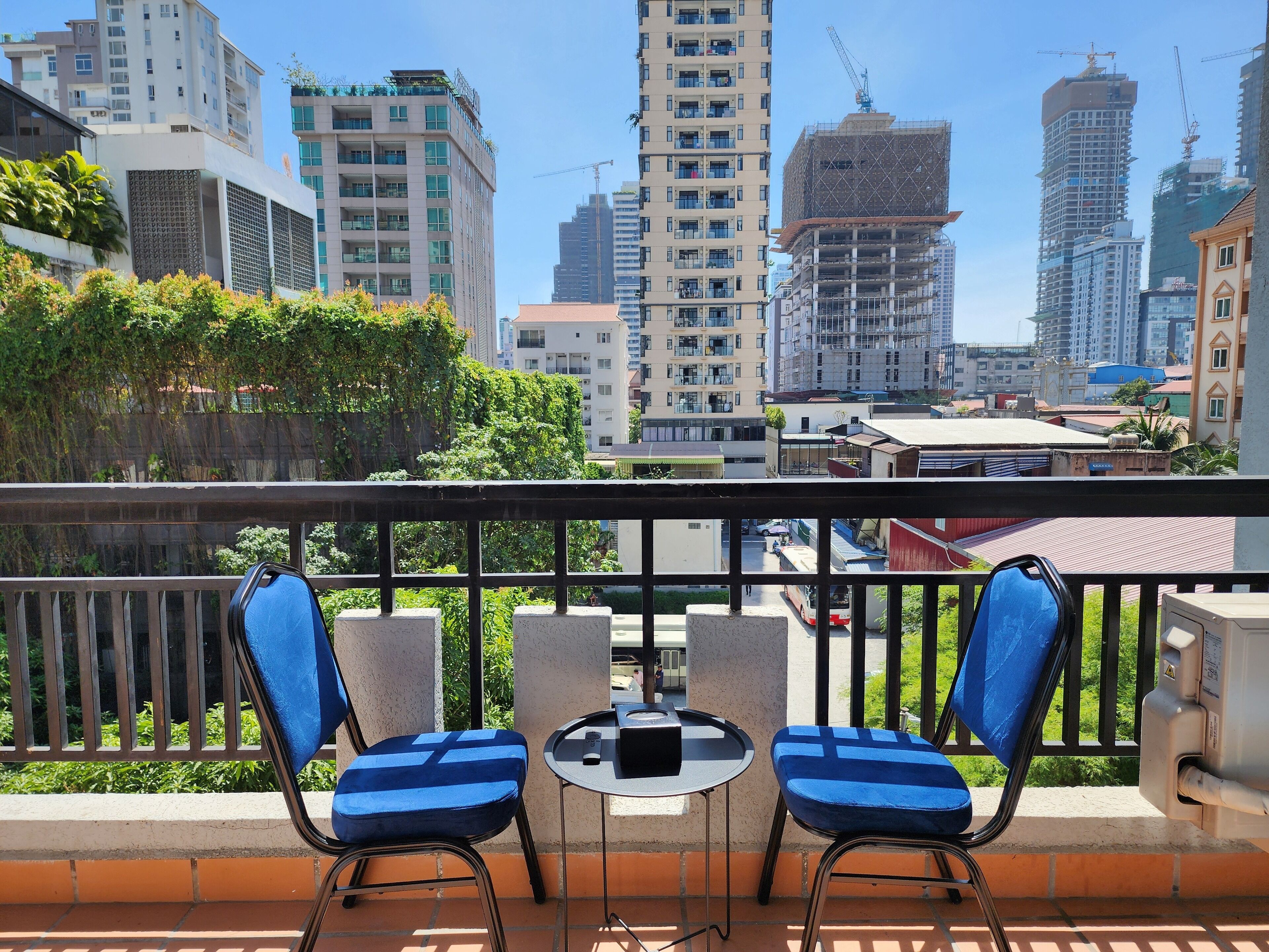 Varanda com duas cadeiras azuis acolchoadas e uma pequena mesa, com vista para um horizonte urbano com diversos edifícios, incluindo um canteiro de obras.