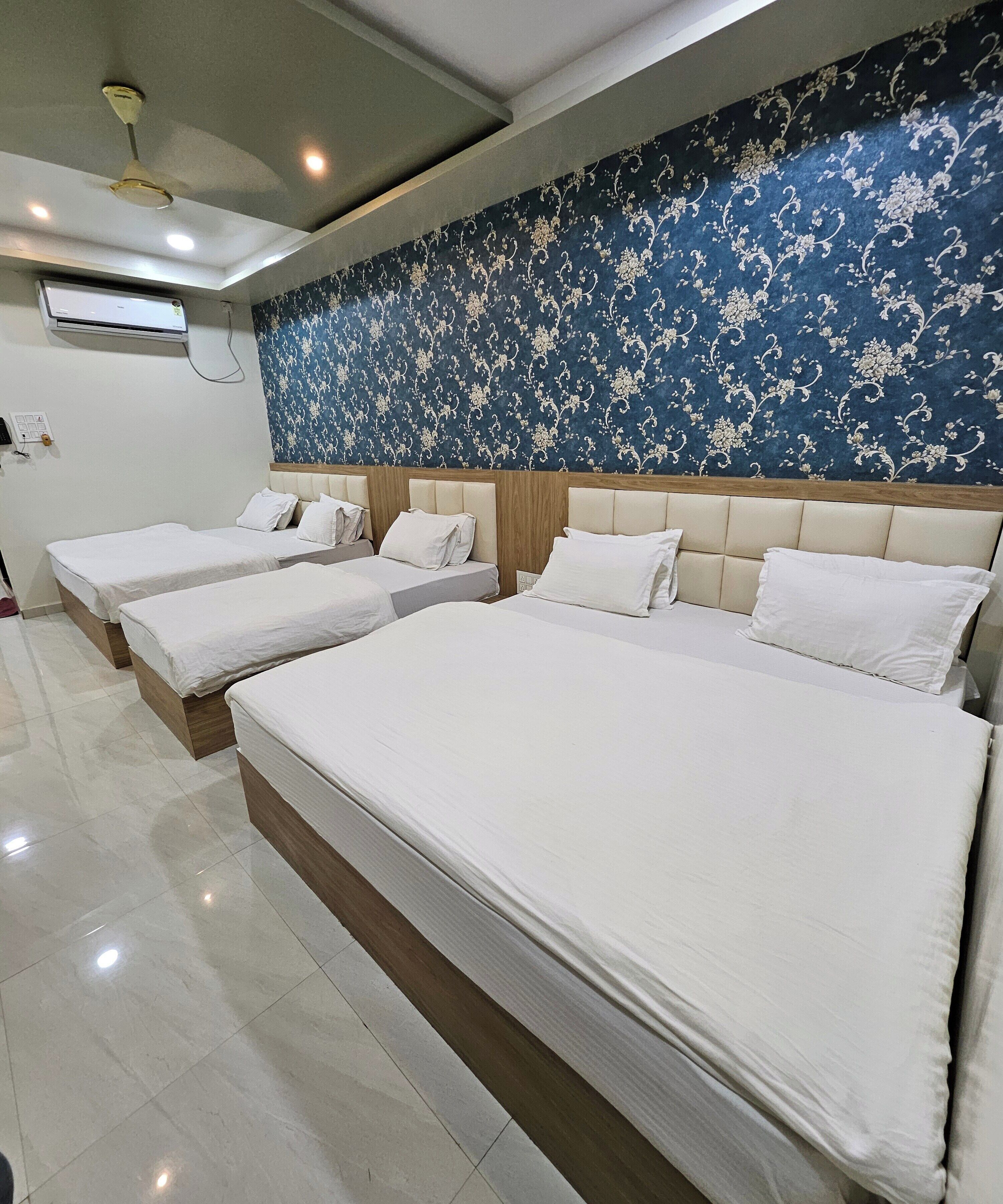 Quarto de hotel com três camas, ventoinha no teto, ar condicionado e parede com padrão floral.