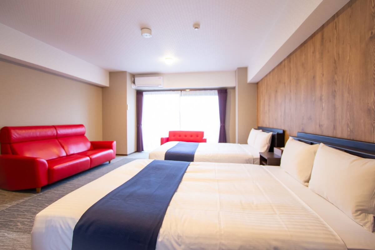 Quarto de hotel com duas camas, um sofá vermelho, uma cabeceira de madeira e uma janela com cortinas.