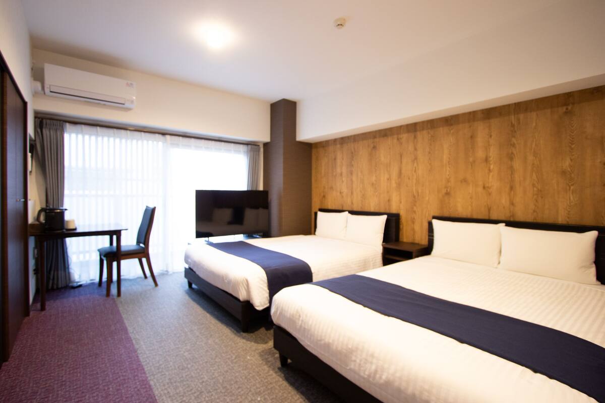Quarto de hotel com duas camas, uma escrivaninha, uma cadeira e uma televisão.