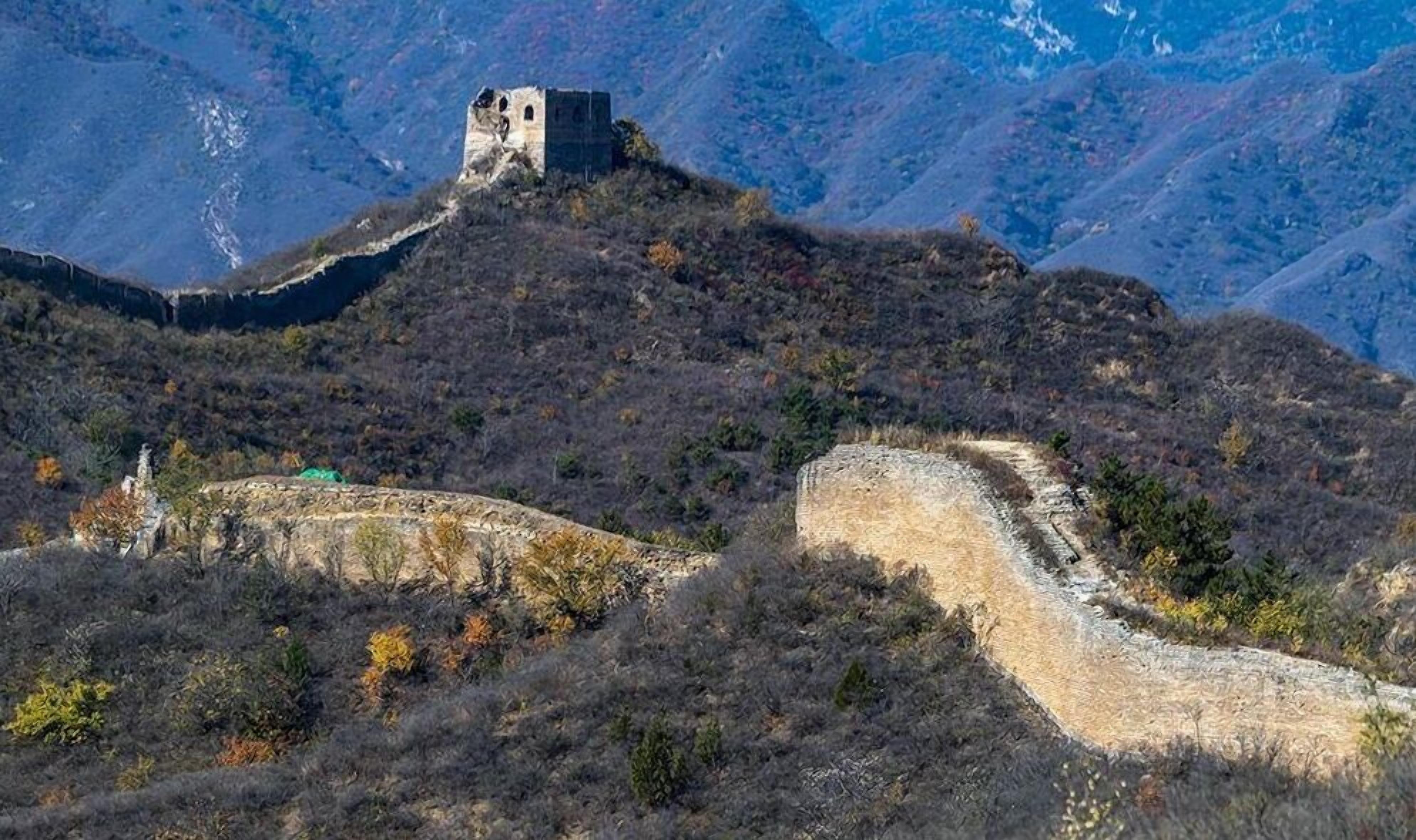 Um muro de pedra histórico com uma torre de vigia numa colina, rodeado por terreno montanhoso.