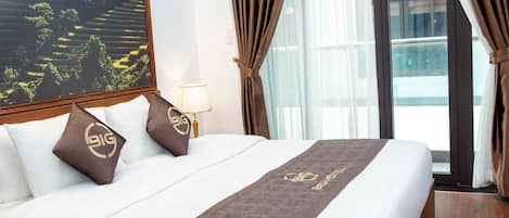 Uma cama bem arrumada com roupa de cama branca e castanha, um quadro grande emoldurado com uma paisagem, e uma janela com cortinas.