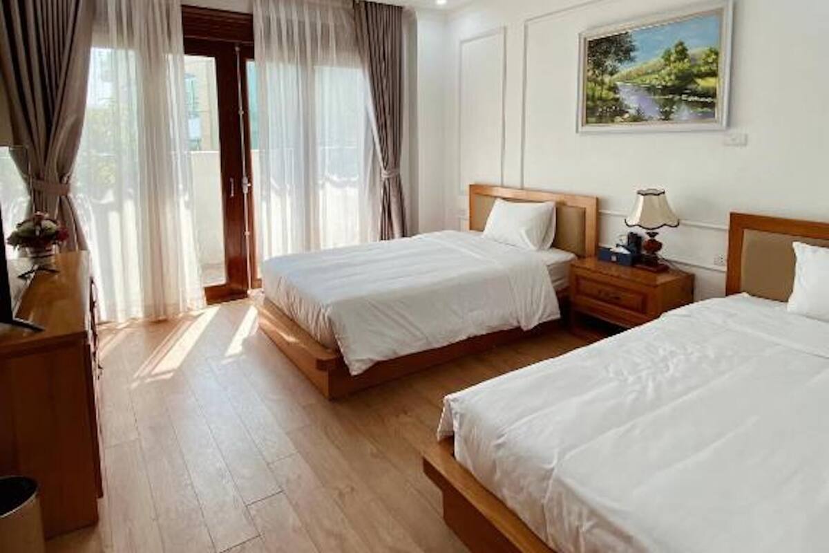 Quarto de hotel com duas camas, soalho de madeira, um quadro na parede e uma janela com cortinas.