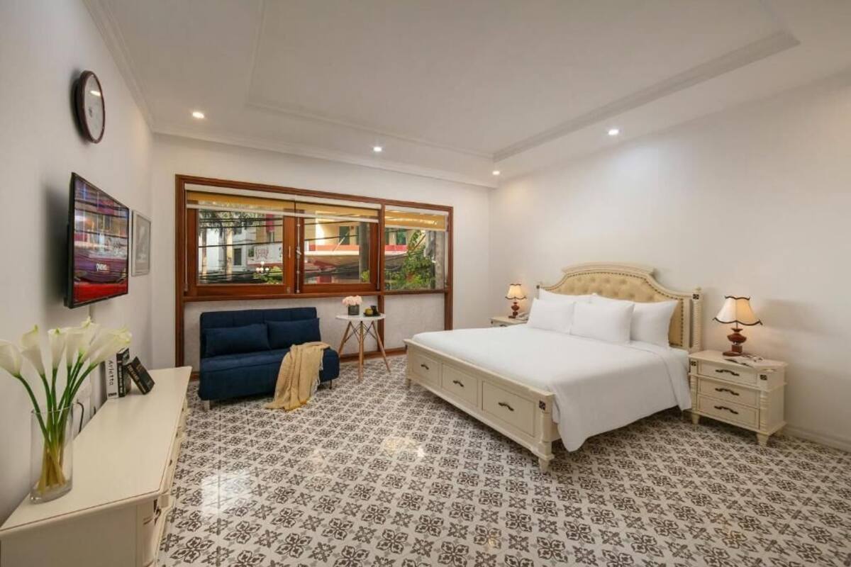 Quarto com uma cama grande, mesa de cabeceira, cômoda, sofá azul, pequena mesa com abajur e uma janela ampla com vista para o exterior.