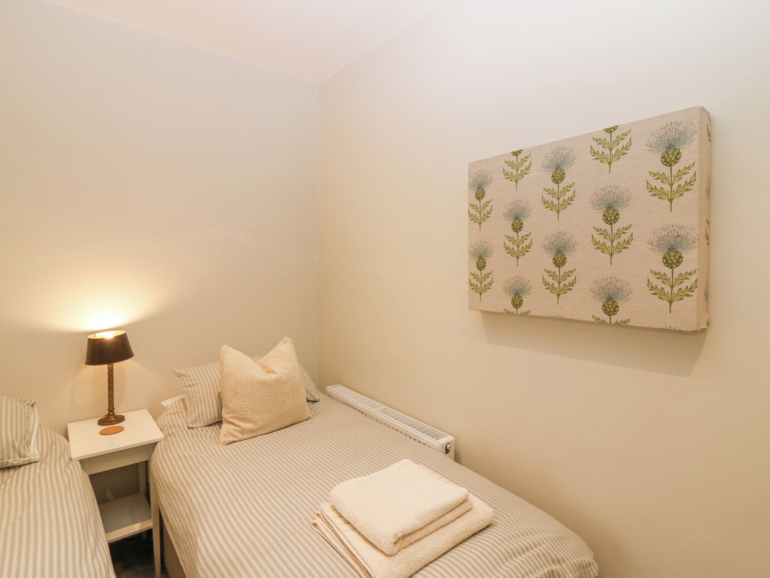 Um quarto pequeno e acolhedor com duas camas, uma mesa de cabeceira, uma luminária e uma obra de arte na parede.
