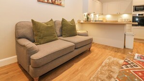 Uma sala de estar moderna com um sofá cinza, almofadas estampadas em verde, piso de madeira, área de cozinha com armários brancos e um quadro de vacas na parede.