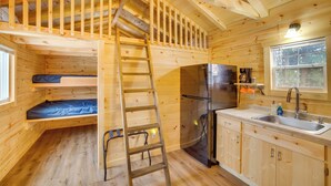 Uma cabana de madeira compacta com quarto no mezanino, cozinha com frigorífico e janela que proporciona luz natural.