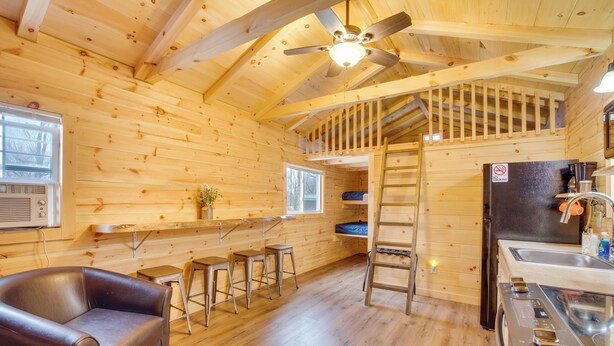 Interior de cabana com decoração em madeira, cozinha compacta, sofá e escada para um andar superior.
