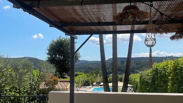 Uma área de estar coberta ao ar livre, vista para a piscina, mesa com bebida e um frasco decorativo.