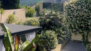 Uma casa com um alpendre de pedra, cercada por vegetação exuberante e uma árvore alta.