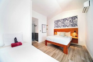 Quarto moderno com cama de madeira, roupa de cama branca, um travesseiro vermelho, ar-condicionado fixado na parede e uma obra de arte emoldurada acima da cama.