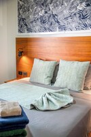 Cama bem arrumada com colcha estampada e almofadas, cabeceira de madeira, luminária na parede e decoração na parede acima da cama.