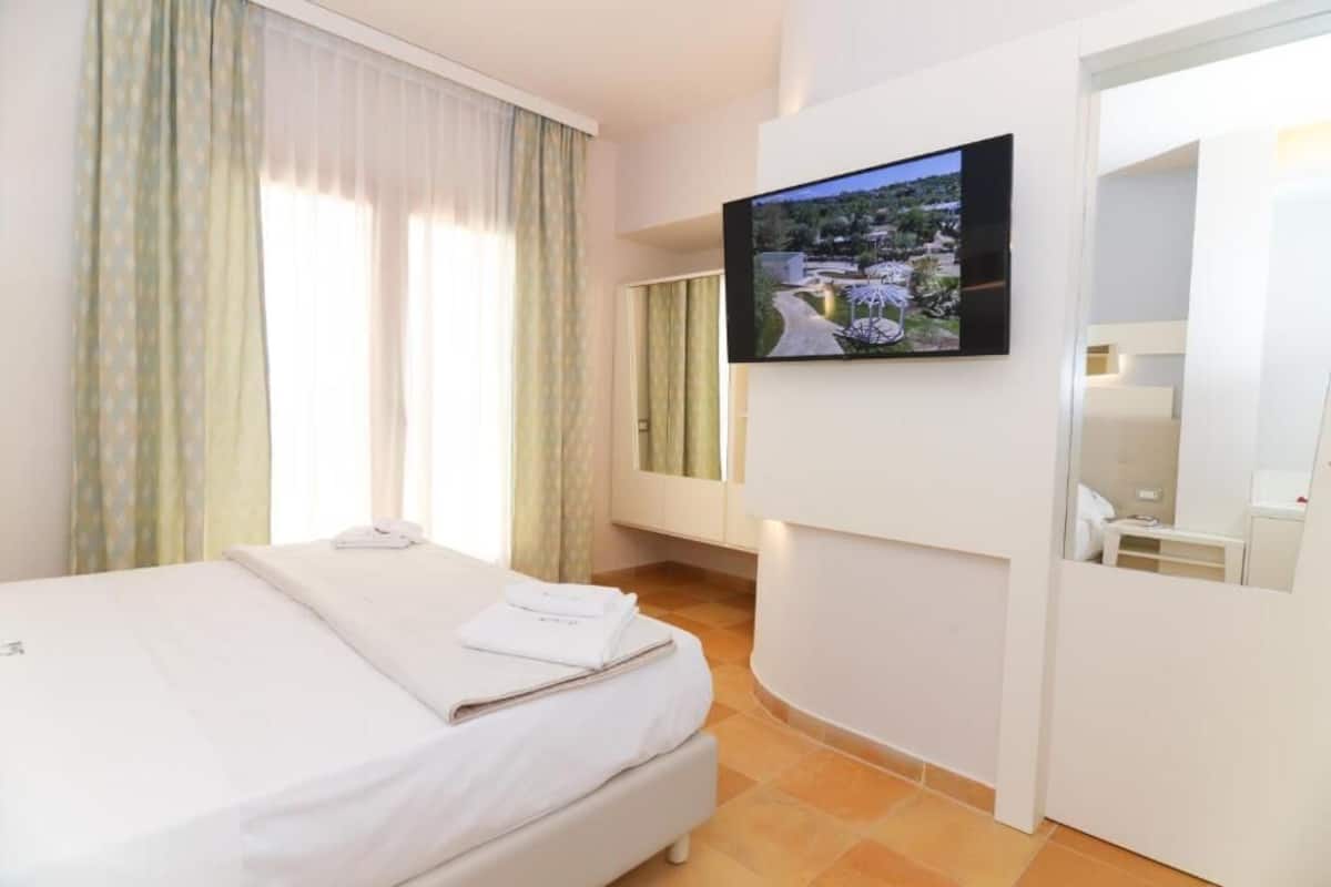 Quarto de hotel com cama, televisão na parede e janela com cortinas.