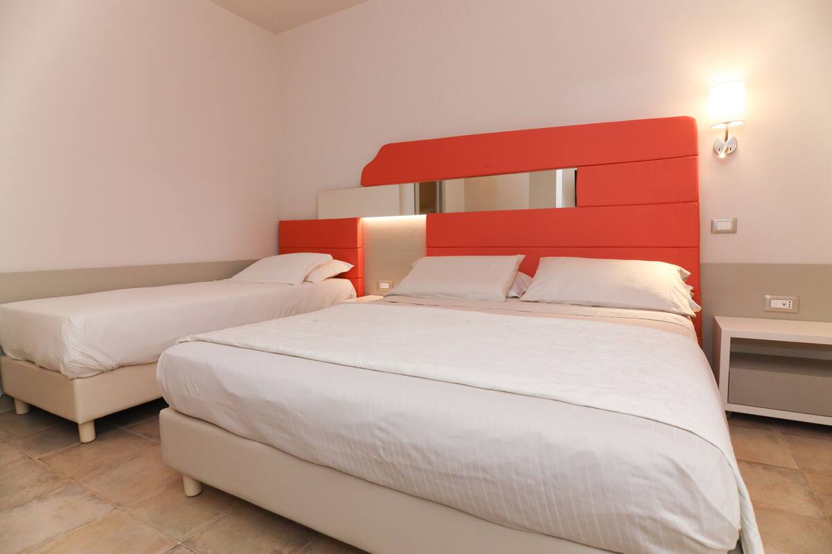 Quarto de hotel com uma cama grande, duas camas menores, uma mesinha de cabeceira e uma luminária fixada na parede.