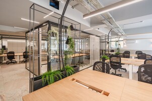 Um espaço de escritório com divisórias de vidro, mesas de madeira, cadeiras de escritório pretas e plantas em vasos.