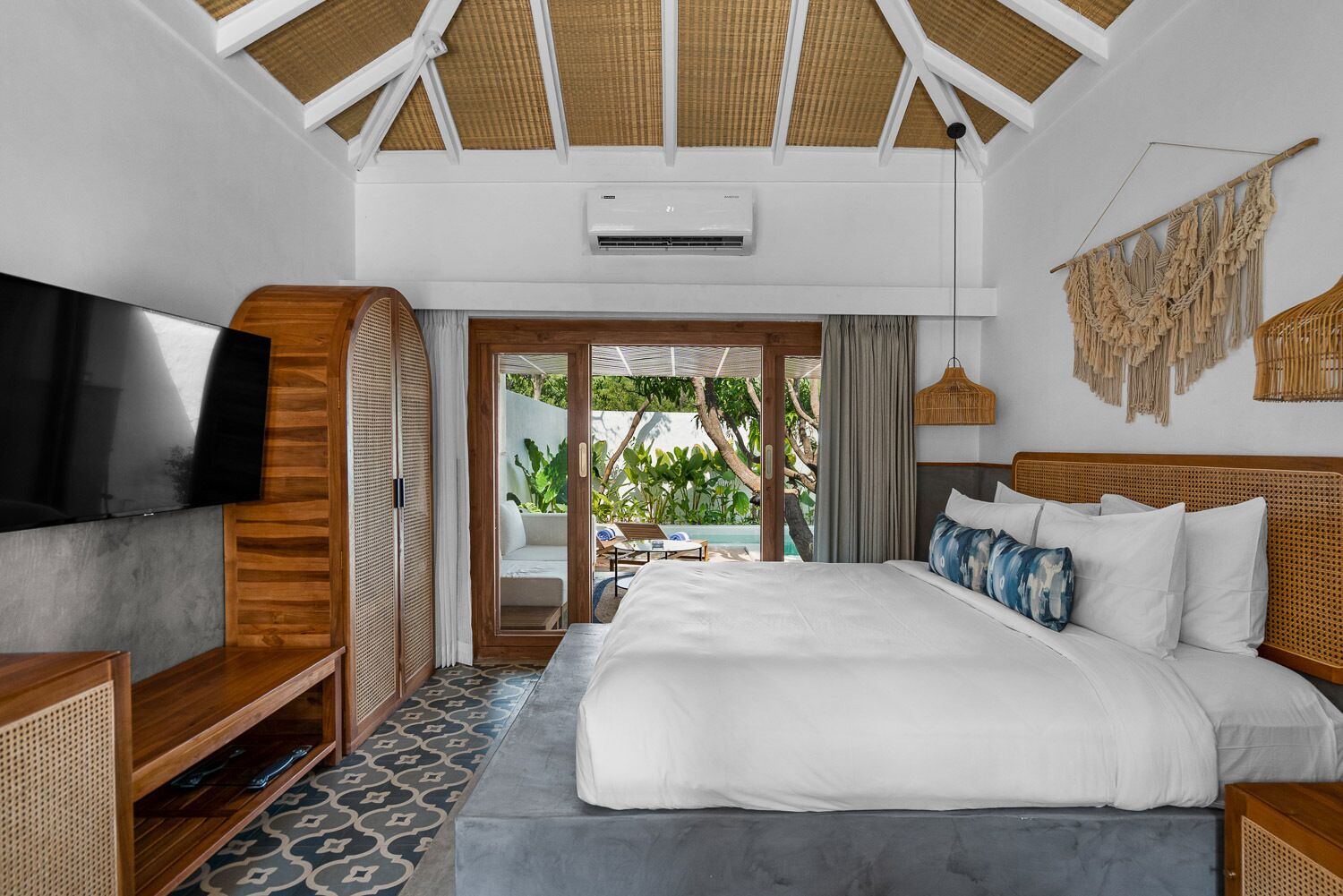 Quarto com uma cama grande, televisão, um banco de madeira e vista para um jardim tropical.
