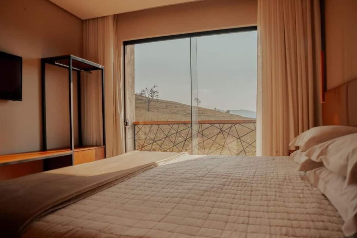 Quarto de hotel com cama, televisão, guarda-roupa e varanda com vista para uma colina.