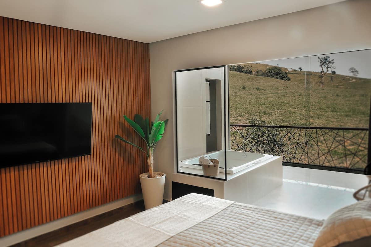 Quarto moderno com televisão fixada em uma parede de painel de madeira, planta em vaso, espelho grande e varanda com vista para uma paisagem gramada.