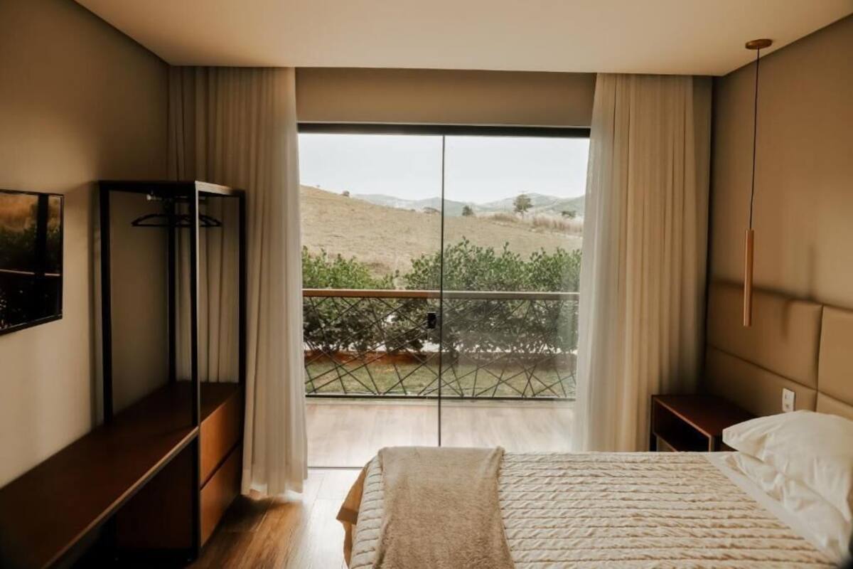 Quarto com uma cama grande, mesa de cabeceira, um banco de madeira, guarda-roupa e vista para uma varanda com grade e vegetação.