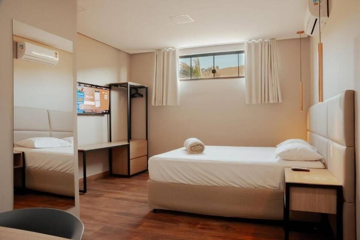 Quarto de hotel com cama, escrivaninha, cadeira e janela com cortinas.