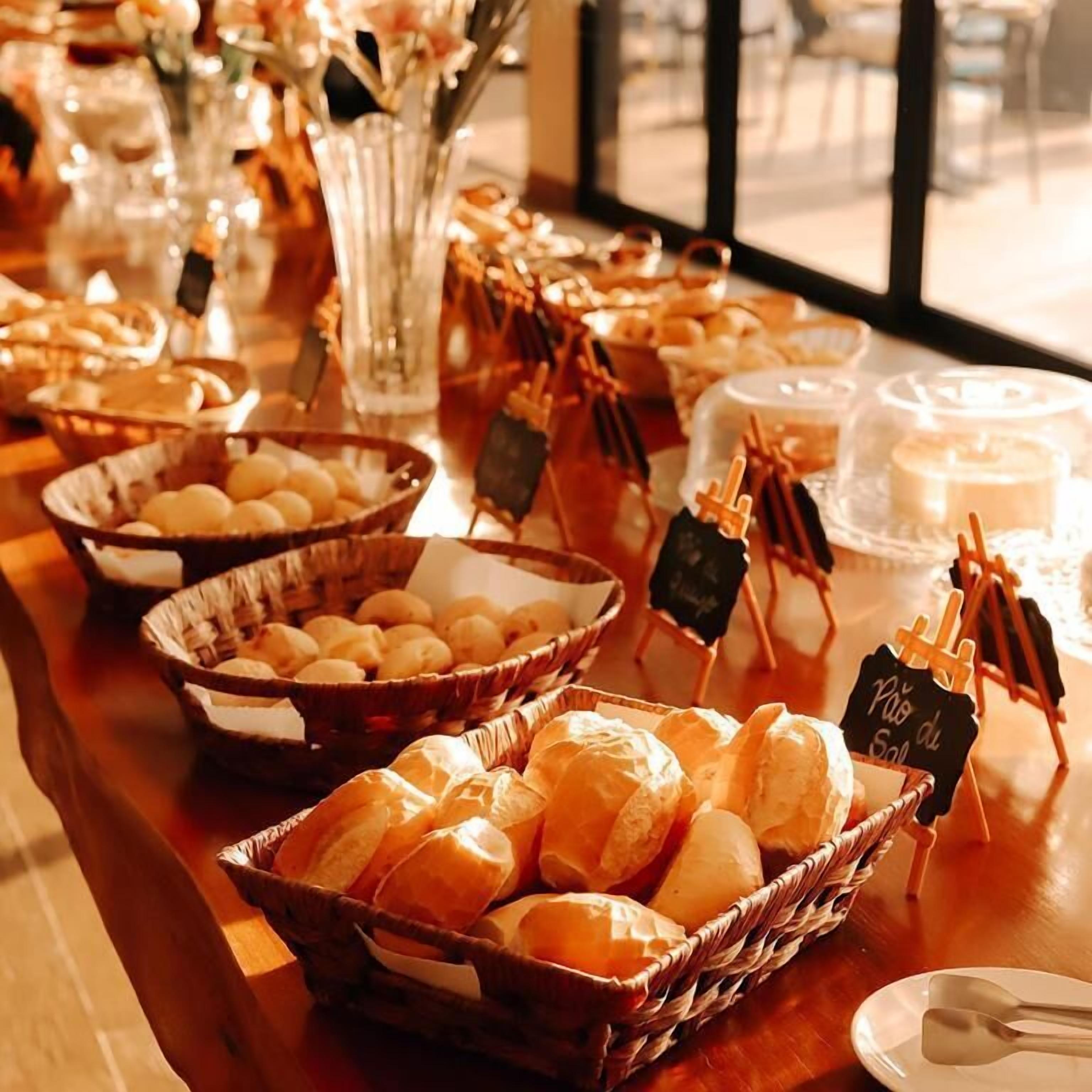 Uma mesa de buffet com diversos alimentos, incluindo cestas de pão e doces, e um castiçal com uma placa.