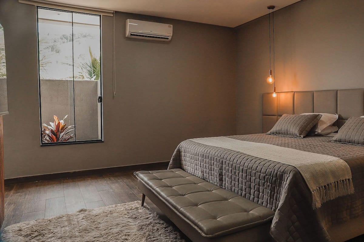 Quarto moderno com uma cama grande, um banco, uma janela com vista e um ar condicionado.