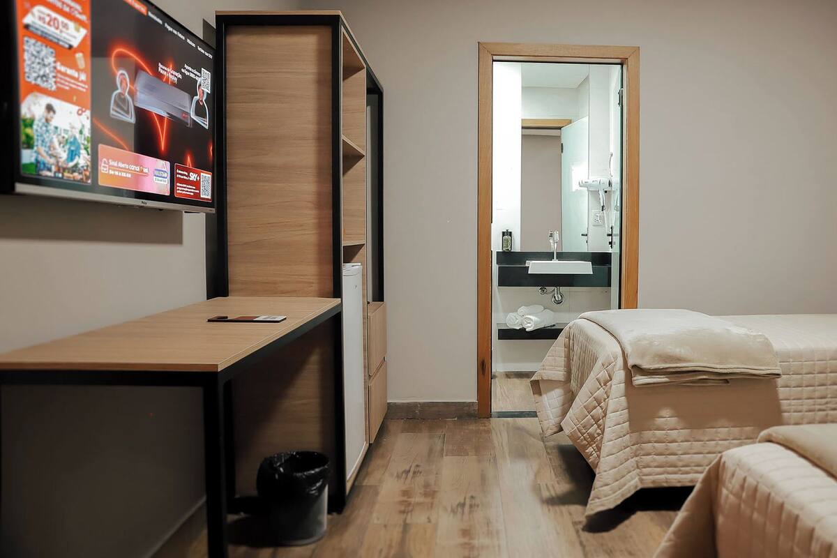 Quarto de hotel com escrivaninha de madeira, cama, banheiro pequeno e televisão fixada na parede.