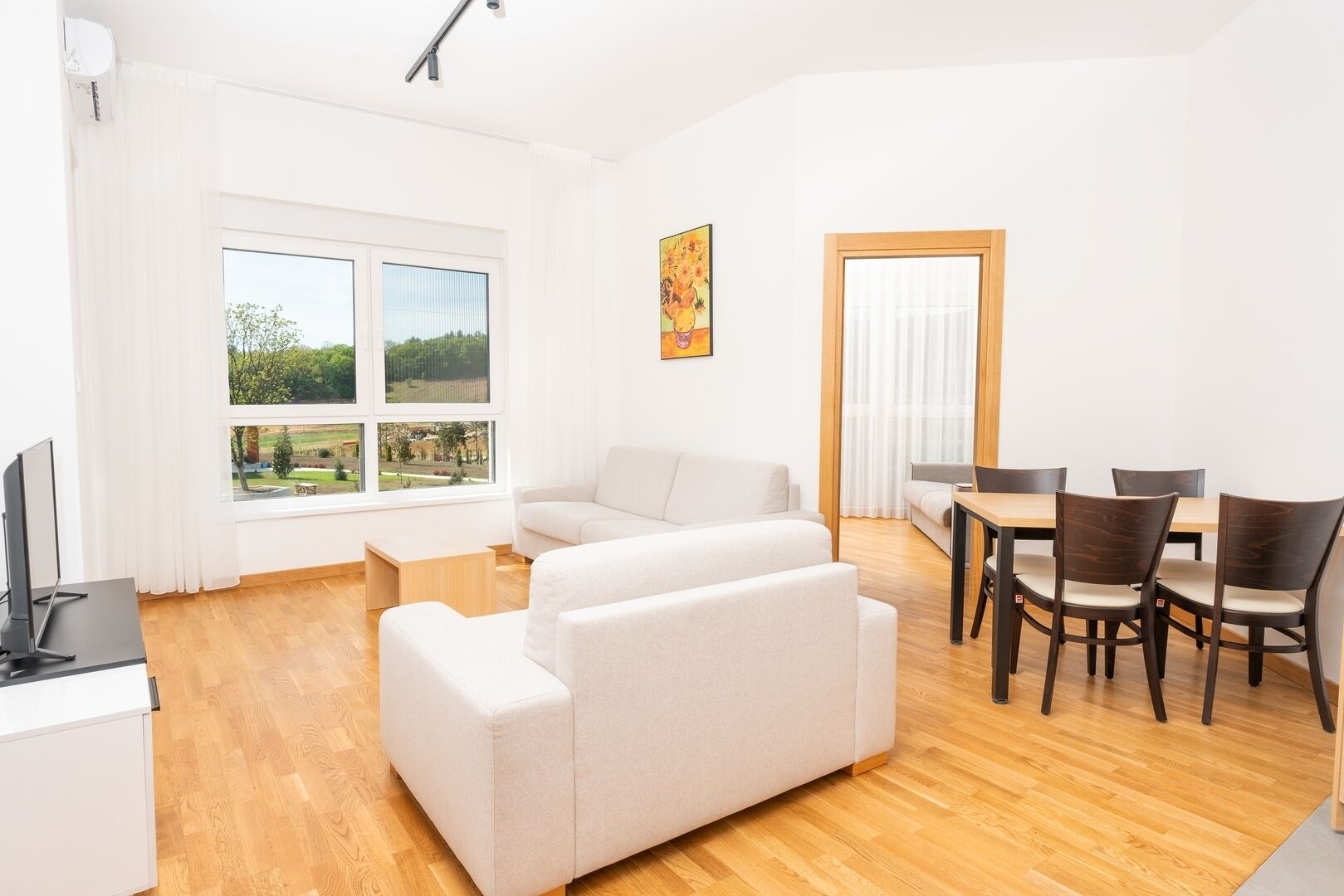 Uma sala de estar moderna com um sofá, uma mesa de jantar com cadeiras, uma televisão e janelas amplas com vista para o verde.