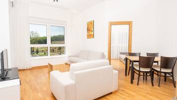 Uma sala de estar moderna com um sofá, uma mesa de jantar com cadeiras, uma televisão e janelas amplas com vista para o verde.