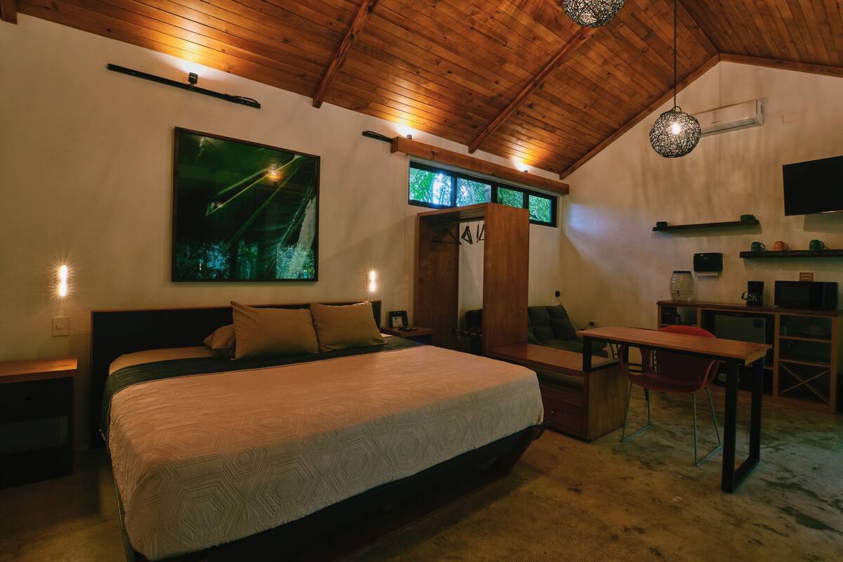 Quarto com uma cama grande, uma escrivaninha e uma cadeira. Há uma televisão na parede e uma janela com vista para árvores.