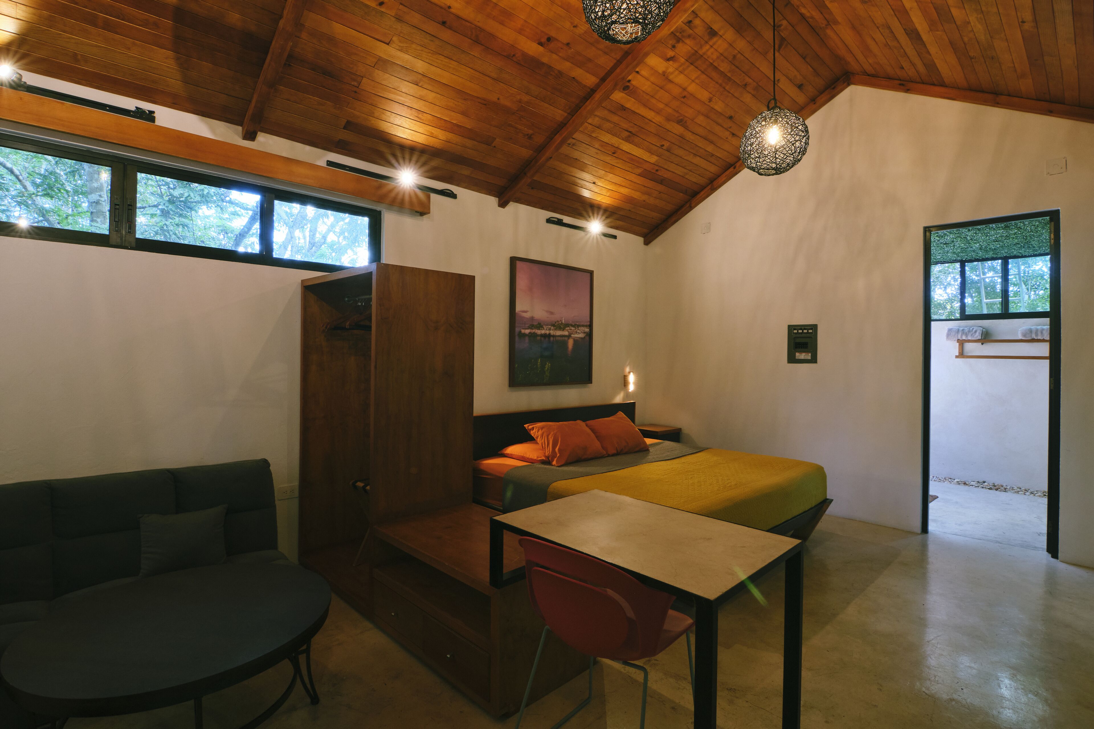 Quarto compacto com cama, sofá, mesa e cadeira. Há janelas e uma porta.