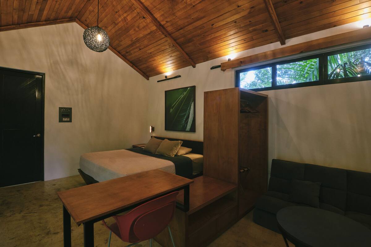 Quarto com cama, escrivaninha, cadeira, sofá e janela com vista para árvores.