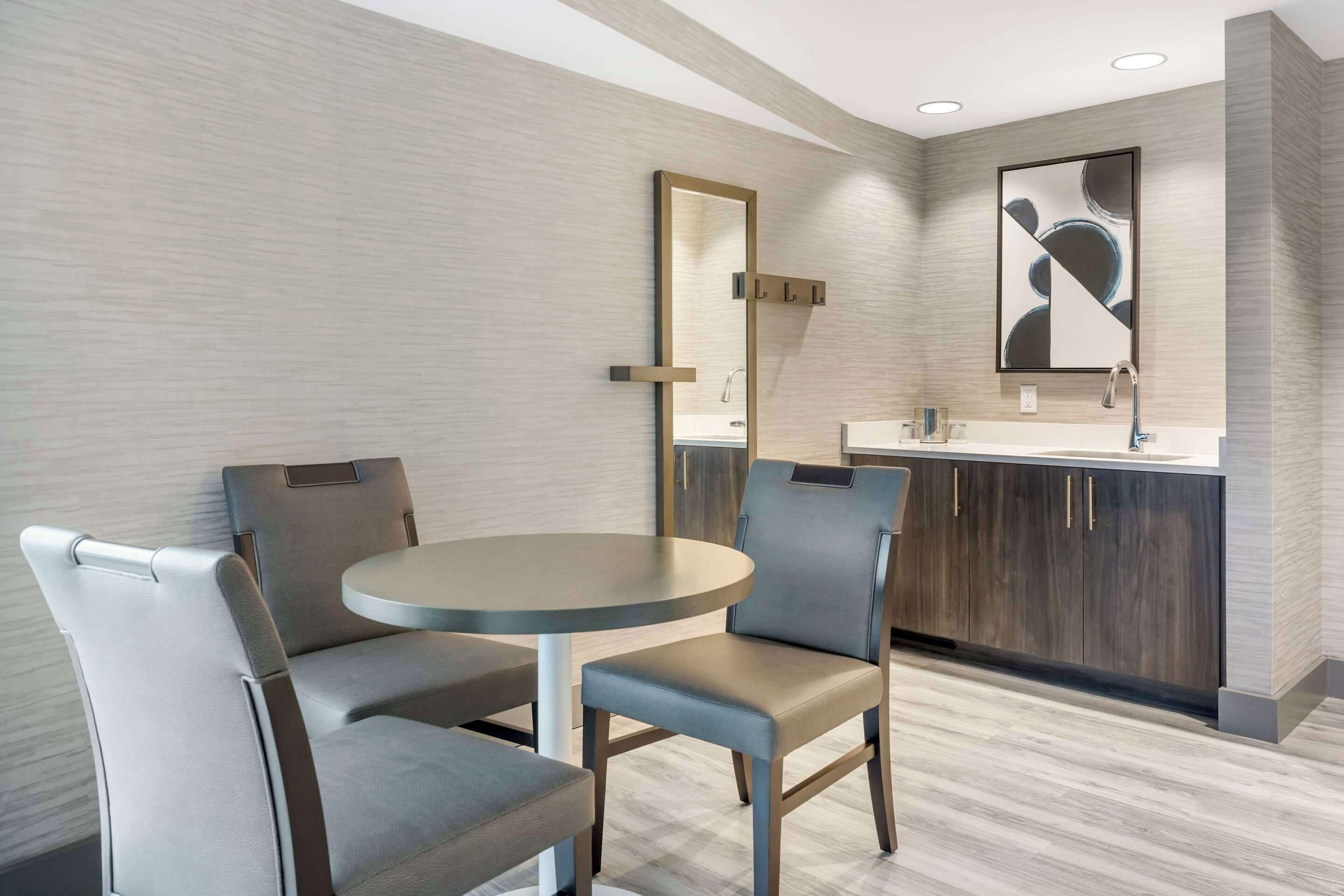 Quarto de hotel moderno com uma mesa de jantar redonda, duas cadeiras cinzas estofadas, um armário de madeira, um espelho grande e uma obra de arte abstrata contemporânea na parede.