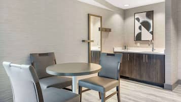 Quarto de hotel moderno com uma mesa de jantar redonda, duas cadeiras cinzas estofadas, um armário de madeira, um espelho grande e uma obra de arte abstrata contemporânea na parede.
