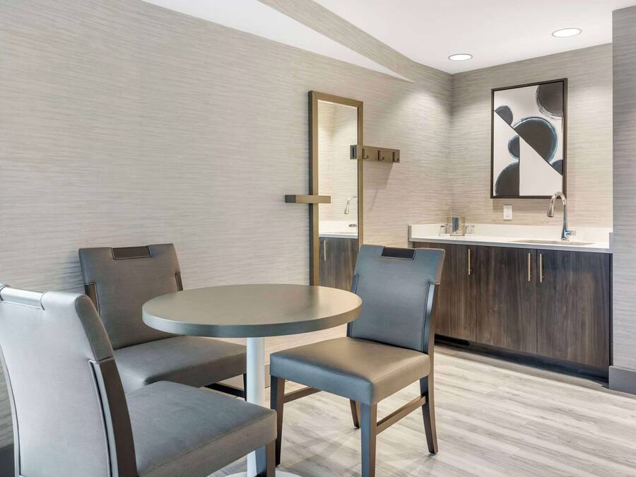 Quarto de hotel moderno com uma mesa de jantar redonda, duas cadeiras cinzas estofadas, um armário de madeira, um espelho grande e uma obra de arte abstrata contemporânea na parede.